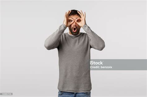 회색 스웨터에 놀랍고 흥분하고 매혹적인 잘 생긴 수염을 가진 남자가 안경이나 쌍안경과 같은 눈 위에 손가락으로 동그라미를 치고 입을 벌리고 즐겁게 말하고 멋진 프로모션을