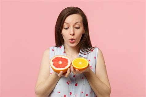 분홍색 파스텔 벽 배경에 고립 된 신선한 잘 익은 자 몽 오렌지 과일의 Halfs를 들고 여름 옷에 놀 랐 젊은 여자 사람들은 생생한 라이프 스타일 휴식 휴가 개념입니다