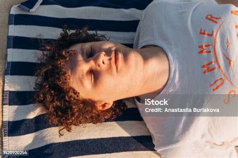 곱슬 머리 눈을 감고 쉬고 휴식을 취하거나 여름이라는 단어가있는 흰색 티셔츠를 입고 자고있는 젊은 백인 남성은 일몰시 해변의 줄무늬 수건에 누워 있습니다 세로 정면 상단보기