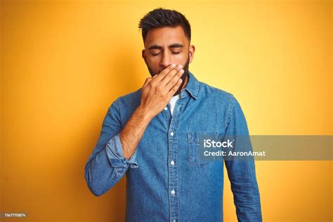 고립 된 노란색 배경 위에 서있는 데님 셔츠를 입은 젊은 인도 남자가 손으로 입을 덮는 피곤한 하품 불안과 졸음 가리기에 대한 스톡 사진 및 기타 이미지 Istock