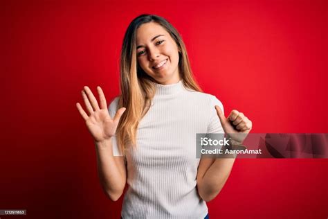 빨간색 배경 위에 캐주얼 한 흰색 티셔츠를 입고 자신감과 행복 미소를 지으면서 손가락 번호 6을 가리키는 파란색 눈을 가진 아름다운 금발 여성 T 셔츠에 대한 스톡 사진 및