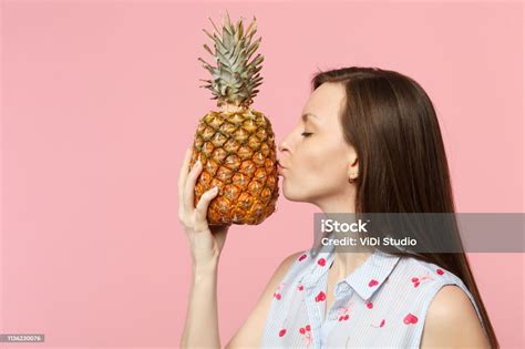 여름 옷에 젊은 여자의 측면 보기 스튜디오에서 분홍색 파스텔 벽 배경에 고립 된 신선한 잘 익은 파인애플 과일 키스를 개최
