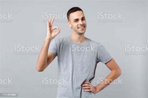 캐주얼에 서 있는 매력적인 젊은 남자의 초상화 카메라를 찾고 확인 제스처 회색 벽 배경에 고립 표시 사람들은 진심으로 감정 라이프 스타일 개념입니다 복사 공간을 모의 합니다
