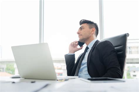 검은 색 양복을 입은 똑똑한 잘생긴 사업가는 사무실의 직장에서 랩톱 컴퓨터에서 작업하는 동안 파트너 및 고객과 비즈니스 거래를 협상하기 위해 휴대 전화를 사용하고 있습니다