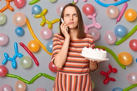 갈색 머리에 줄무늬 드레스를 입고 케이크를 들고 휴대폰으로 이야기하는 슬픈 여자는 화려한 풍선으로 장식된 회색 벽에 서서 올려다본다 연결에 대한 스톡 사진 및 기타 이미지