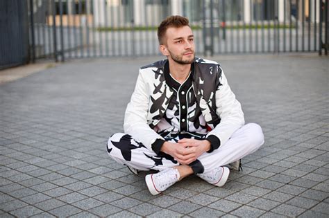 장난기 넘치는 표정으로 흰색과 검은 색 양복을 입은 잘 생긴 남자 트렌디 한 양복을 입은 그루터기와 모델 헤어 스타일을 가진 남자가 포장 도로에 앉아 있습니다 25 29세에