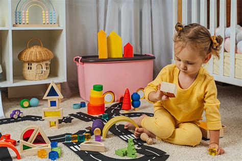 바닥에 앉아있는 어린 소녀는 놀이방에서 장난감 나무 블록 자동차와 도로를 가지고 놀고 있습니다 현대 보육원에서 아기와 유아를위한 교육 게임 몬테소리 교육에 대한 스톡 사진