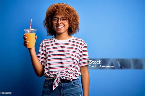 젊은 아프리카계 미국인 여성 곱슬 머리 와 곱슬 머리 를 입고 모자 를 마시는 오렌지 주스 와 행복한 얼굴 서서 치아를 보여주는 자신감 미소와 미소 마시기에 대한 스톡 사진