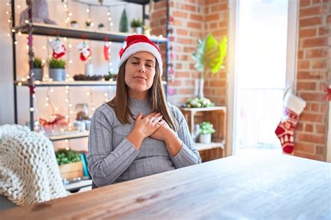 집에서 식탁에 앉아 있는 크리스마스 모자를 쓰고 있는 젊은 아름다운 여성은 눈을 감고 얼굴에 감사하는 제스처를 입고 가슴에 손을