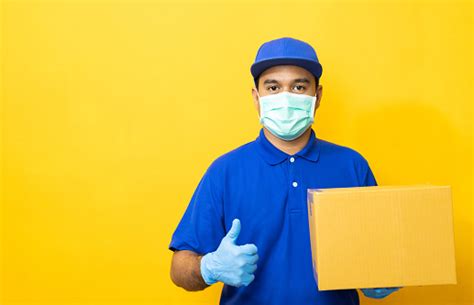 배달 남자 블루 유니폼 고무 장갑을 착용 하 고 노란색 배경에 소포 판지 상자를 들고 마스크 배달부에 대한 스톡 사진 및 기타 이미지 Istock