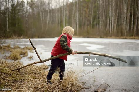 이른 봄날 숲 호수 기슭에서 큰 가지를 노는 어린 소년 호수 표면은 여전히 얼음 아래에 있습니다 아이는 막대기로 얼음을 깨려고합니다 어린이를 위한 야외 활동 6 7 살에