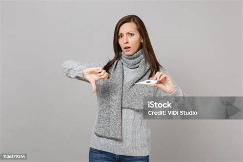 아래로 엄지손가락을 보여주는 회색 스웨터 스카프에 젊은 여자를 의아해 회색 벽 배경에 고립 된 유지 온도계 건강 한 라이프 스타일 아픈 질병 치료 추운 계절 개념입니다 복사
