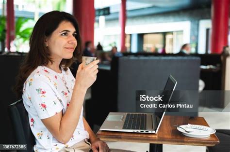 긍정적 인 표정으로 웃는 여성을보고 거리를 바라 보는 커피 한 잔을 들고있는 사이드 뷰 컴퓨터 노트북으로 카페 테이블에 앉아있는