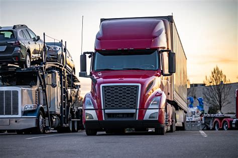 드라이 밴 세미 트레일러가 있는 부르고뉴 빅 리그 세미 트럭이 황혼의 일몰 시간에 다른 세미 트럭과 함께 산업 주차장에 서 있습니다 화물 자동차 휴게소에 대한 스톡 사진 및