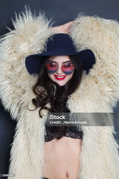 밝은 메이크업과 긴 웨이브 머리를 가진 행복 한 젊은 성인 여성 패션 모델 파란색 모자와 선글라스를 착용 푸른 스튜디오 벽 배경에 포즈를 취하고 있습니다 18 19세에 대한