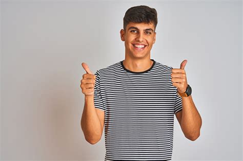 해군 줄무늬 티셔츠를 입고 고립 된 흰색 배경 성공 표지판 위에 서있는 젊은 인도 남자는 손으로 긍정적 인 제스처를하고 엄지 손가락으로 미소와 행복을 하고 있습니다 쾌활한