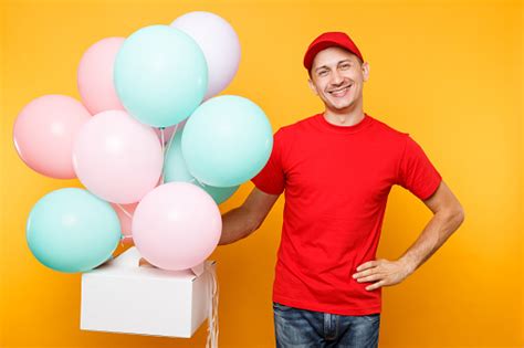음식 주문 케이크 상자 노란색 배경에 절연을 주는 남자 남성 직원 택배 빨간 모자 티셔츠 보류 다채로운 공기 풍선에 빈 골 판지 상자에서 디저트 배달 서비스 개념입니다