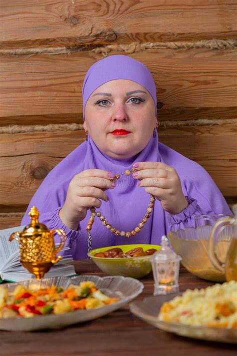 A Muslim Woman In A Lilac Hijab Is Fingering Her Rosary At The Table Praying For The End Of