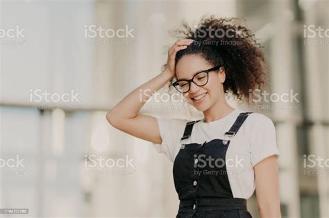 선명한 머리와 아름 다운 어두운 피부 여성 모델 아래를 보고 캐주얼 흰색 티셔츠 검은 바지 여가 시간을 즐긴다 야외 산책 좋은 소식에 만족 좋은 감정과 사람들 아프로 머리에