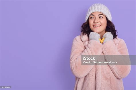 따뜻한 모피 코트와 모자를 쓰고 파스텔 라일락 색상 배경에 고립 된 옆으로 찾고 긍정적 인 감정을 가지고 프로모션공간을 복사 미소 꽤 쾌활한 젊은 여성 20 24세에 대한