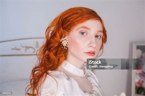 그녀의 귀에 흰색 진주 귀걸이와 흰색 빈티지 웨딩 드레스에 긴 붉은 곱슬 머리를 가진 여자의 초상화 창백한 피부 파란 눈 침실에