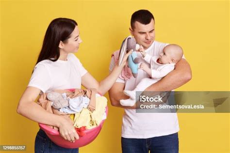 노란 벽에 맞서 사진을 찍고 있는 세 명의 사랑스러운 젊은 가족 엄마가 육지를 하고 더러운 옷으로 가득 찬 분지를 들고 아기를 위로하려고 손에 아기를 안고 있는 아버지