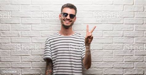 젊은 성인 남자 백색 벽돌 벽 승리 표시를 하 고 카메라에 윙크 하는 행복 한 얼굴로 웃 고 서 선글라스를 착용 번호 2입니다 긍정적인 감정 표현에 대한 스톡 사진 및 기타