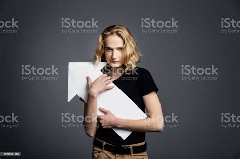 긴 금발 머리와 미소 얼굴을 가진 잘 생긴 젊은 남자가 회색 배경에 오른쪽을 가리키는 흰색 화살표를 포옹 20 24세에 대한 스톡 사진 및 기타 이미지 Istock
