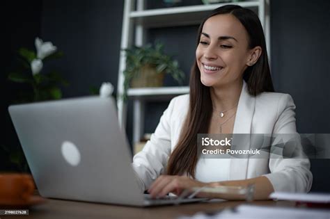 흰색 블레이저를 입은 전문직 여성이 현대적인 사무실 공간의 책상에서 노트북으로 작업하면서 미소를 짓고 있습니다 이 이미지는 생산성 자신감 긍정적인 일과 삶의 균형을