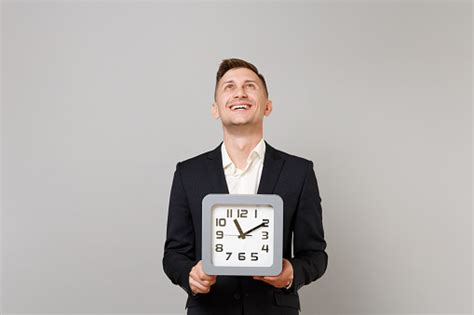 스튜디오에서 회색 벽 배경에 고립 된 사각형 시계를 들고 찾고 고전적인 검은 양복 흰색 셔츠에서 젊은 비즈니스 남자를 웃는 업적 경력 자산 사업 개념 복사 공간을 모의