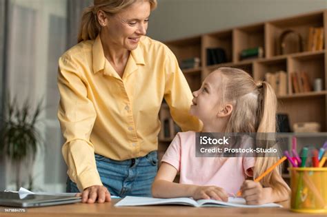 어린 딸이 숙제를 하고 있는지 확인하는 돌보는 어머니 쾌활한 엄마 식탁에 앉아 있는 귀여운 여자 아이 2명에 대한 스톡 사진 및 기타 이미지 Istock