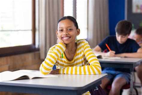 Smiling Girl Sitting At Desk With Open Book In School Classroom Stock