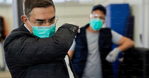 자동차 작업장에서 보호용 안면 마스크를 착용한 상태에서 팔꿈치에 기침을 하는 중년 검사관 배경에 작업자가 있습니다 사진 Unsplash의 세르비아 이미지