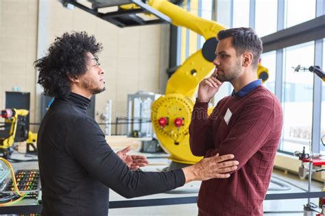 Engineers Discussing Robotics Technology In Industrial Setting With Large Yellow Robotic Arm In