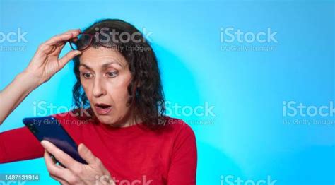 충격과 놀란 성숙한 성인 갈색 머리 백인 여성 빨간 스웨터를 입은 스마트폰 확인 파스텔 블루 배경 위에 복사 공간 Stock Market Crash에 대한 스톡 사진 및