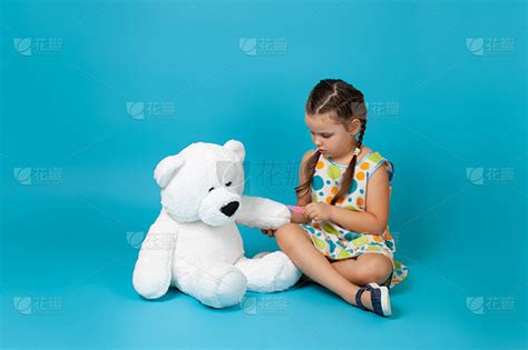 一个女孩盘腿坐在地板上，用镊子从一只白色泰迪熊的爪子里拔出一根刺