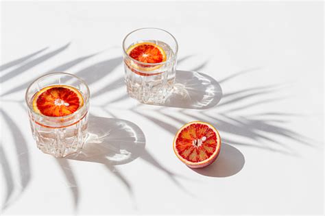 반쯤 피 묻은 붉은 오렌지와 야자수 잎 그림자가 있는 흰색 배경에 칵테일이 있는 두 잔으로 만든 창의적인 구성 여름 다과와 파티 컨셉 미니멀한 스타일 그림자에 대한 스톡