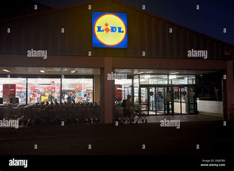 Outside A Lidl Retail Store At Night With Inside Lit Up And Silhouettes Of Trolleys And Customer