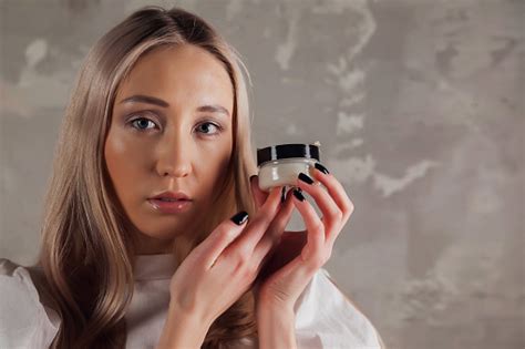 햇빛 그림자에 크림 병 얼굴 피부를 들고 카메라를 보고 있는 귀여운 여자 질감이 있는 벽 배경에 화장품 마사지 크림을 바르고 있는 사랑스러운 아가씨 미용 자기 관리 개념