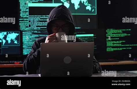 Asian Male Hacker In A Hood Drinking Coffee While Works On A Laptop