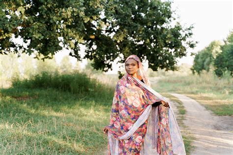 녹색 나무 근처에서 히잡을 입은 매력적인 무슬림 흑인 소녀 녹색 배경에 국가 다채로운 드레스를 입고 아프리카 여성 히잡이라는 스카프를 입고 젊은 흑인 소녀의 라이프 스타일