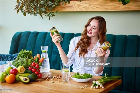 배경에 녹색 꽃과 테이블에 신선한 과일과 야채와 아름다운 인테리어에 스무디와 신선한 물을 마시는 젊은 여성 건강한 식생활 개념 비건 식사 및 해독 메뉴 디톡스에 대한 스톡