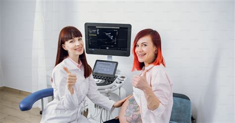 밝은 빨간 머리와 배에 문신을 한 임산부가 임신 검진을 위해 여성 친화적 인 의사를 방문합니다 여성 조산사와 그녀의 임신한 환자가 웃으며 엄지손가락을 치켜들고 있다 사진