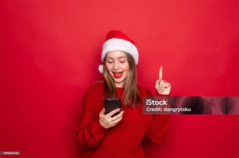 빨간 배경에 전화를 들고 위로 손과 손가락으로 행복 하게 가리키는 크리스마스 모자를 입고 젊은 여자 12월에 대한 스톡 사진 및 기타 이미지 Istock