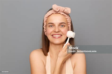 회색 배경 위에 고립되어 서 있고 씻기 위해 브러시를 사용하고 아침 일상적인 절차를 수행하고 행복한 표정으로 카메라를 보고 피부 관리를 즐기는 행복한 여성의 실내 사진 30