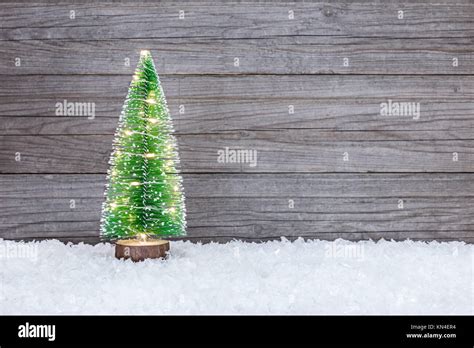 Small Artificial Green Christmas Tree Illuminated With Holiday Lights On Snow Against Wooden
