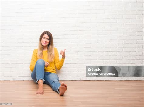 젊은 성인 여자 생기고 엄지 측면 가리키는 행복 한 얼굴로 웃 고 흰색 벽돌 벽을 통해 바닥에 앉아 가리키기에 대한 스톡 사진 및 기타 이미지 Istock