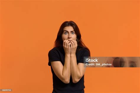 그것은 매우 무서운 검은 색 티셔츠를 입은 긴장한 백인 여성은 손톱을 물고 카메라를 걱정하는 표정으로 보이며 나쁜 것을 기다리며 겁에 질린 표정을 짓고 있습니다 주황색 벽