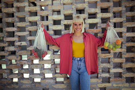 청바지와 넓은 셔츠를 입은 웃는 여성은 야채와 과일이 든 재사용 가능한 가방을 들고 있습니다 25 29세에 대한 스톡 사진 및