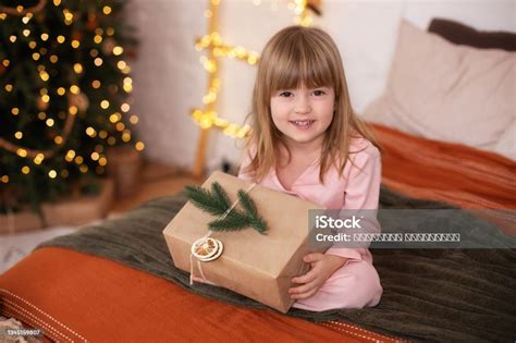 아이는 빛나는 조명 화환과 크리스마스 트리 근처 집에서 새해 선물을 엽니 다 아이는 집에서 아이 침실에서 침대에서 자고 잠옷을